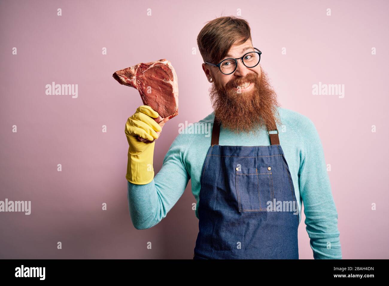 Redhead Irish butcher man with beard holding raw beef steak over pink ...