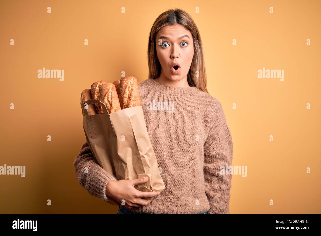 Young beautiful woman holding paper bag with bread over isolated yellow ...