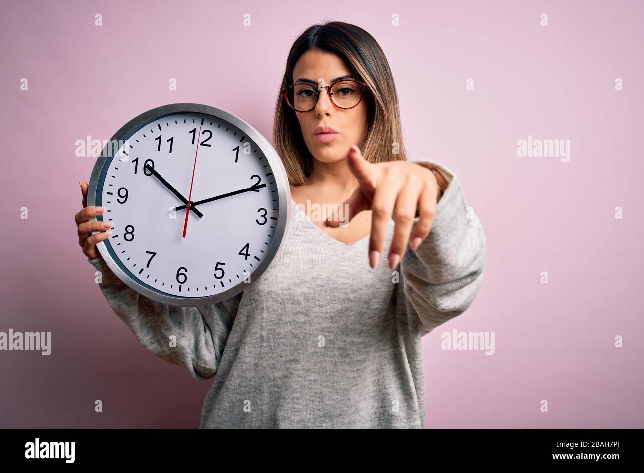Young beautiful brunette woman wearing glasses doing countdown using big clock pointing with ...