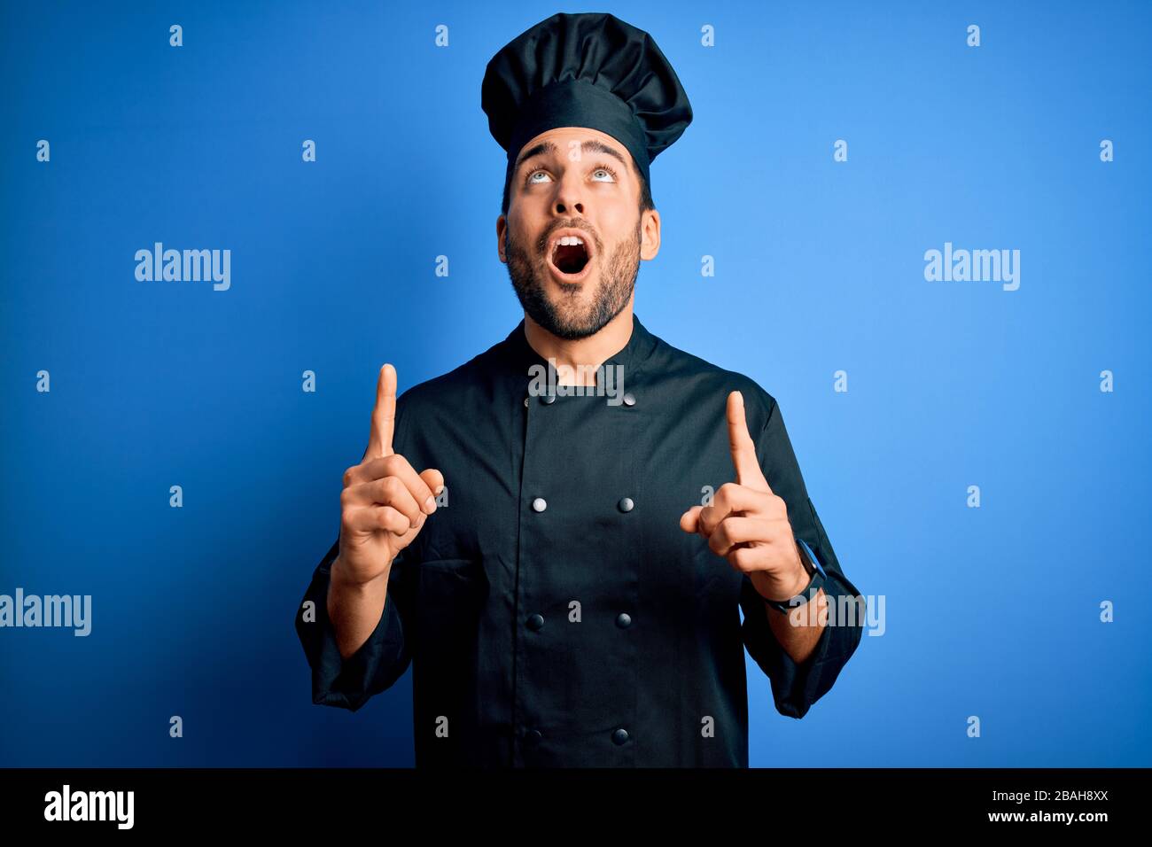 Young handsome chef man with beard wearing cooker uniform and hat over ...