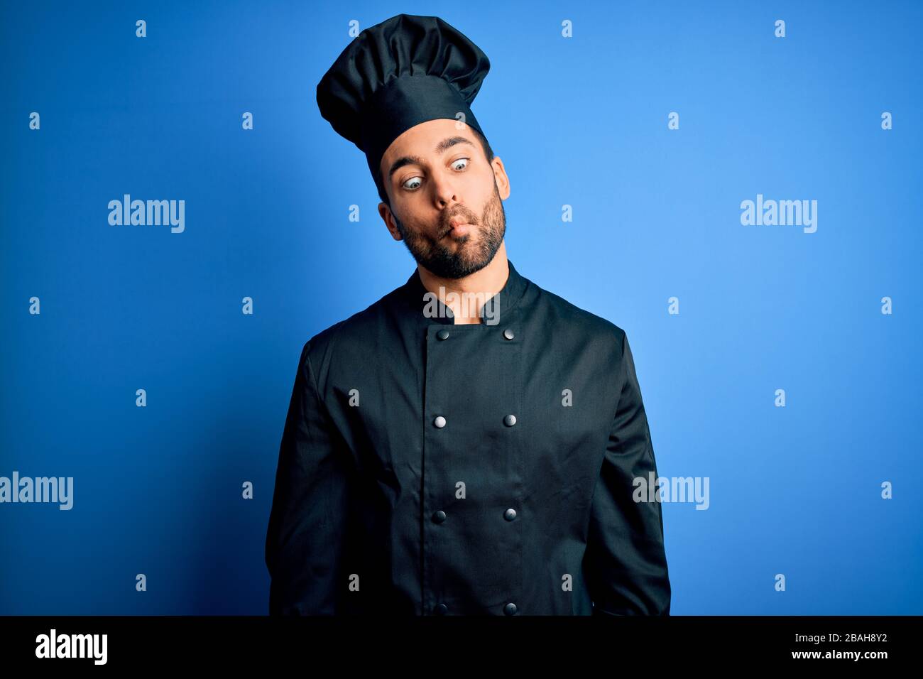 Young handsome chef man with beard wearing cooker uniform and hat over ...