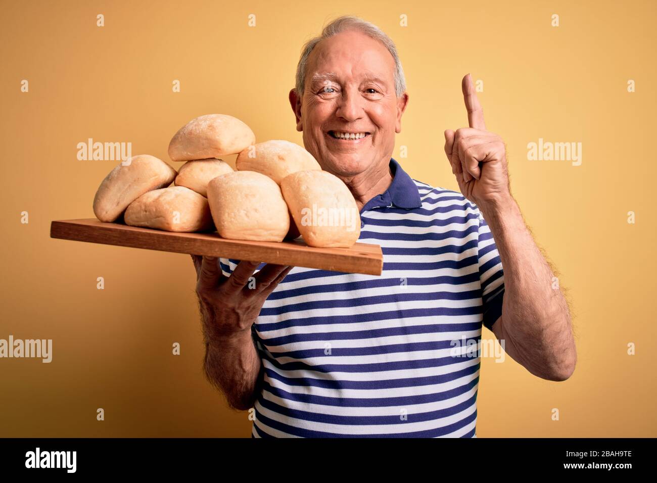 Senior grey haired baker man holding fresh homemade bread over yellow ...