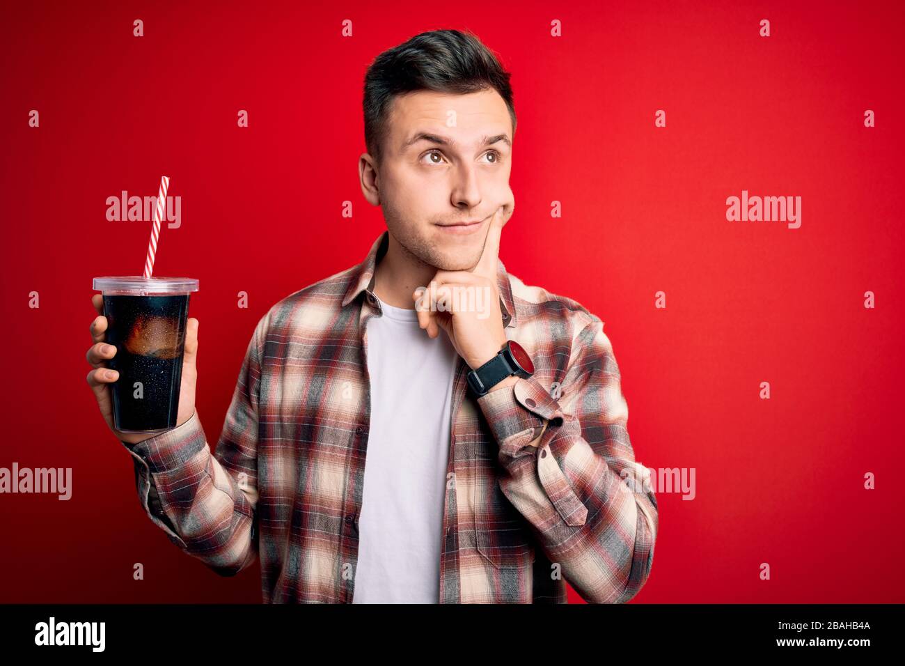 Young handsome caucasian man drinking a fresh soda refreshment over red ...