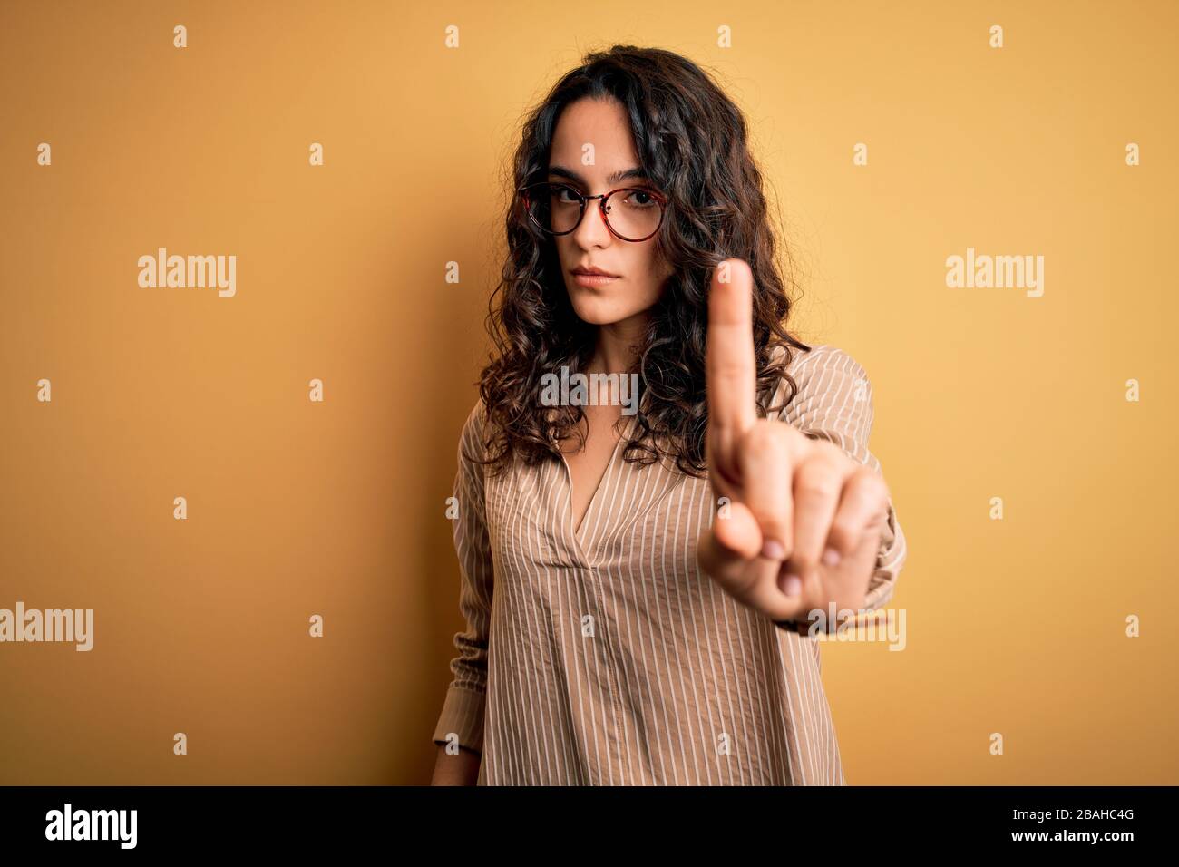 Beautiful woman with curly hair wearing striped shirt and glasses over yellow background ...