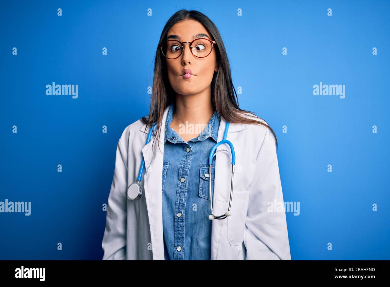 Young beautiful doctor woman wearing stethoscope and glasses over blue background making fish ...