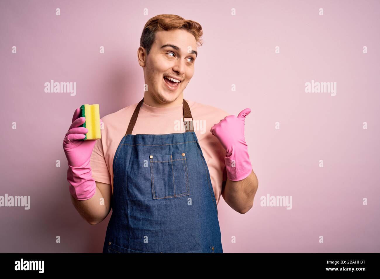 Handsome Redhead Man Doing Housework Wearing Apron And Gloves Using Cleaner Scourer Pointing