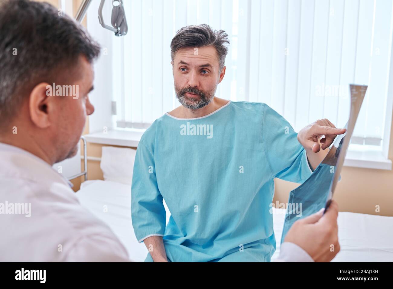 Mature bearded patient in hospital gown sitting on bed and pointing at ...