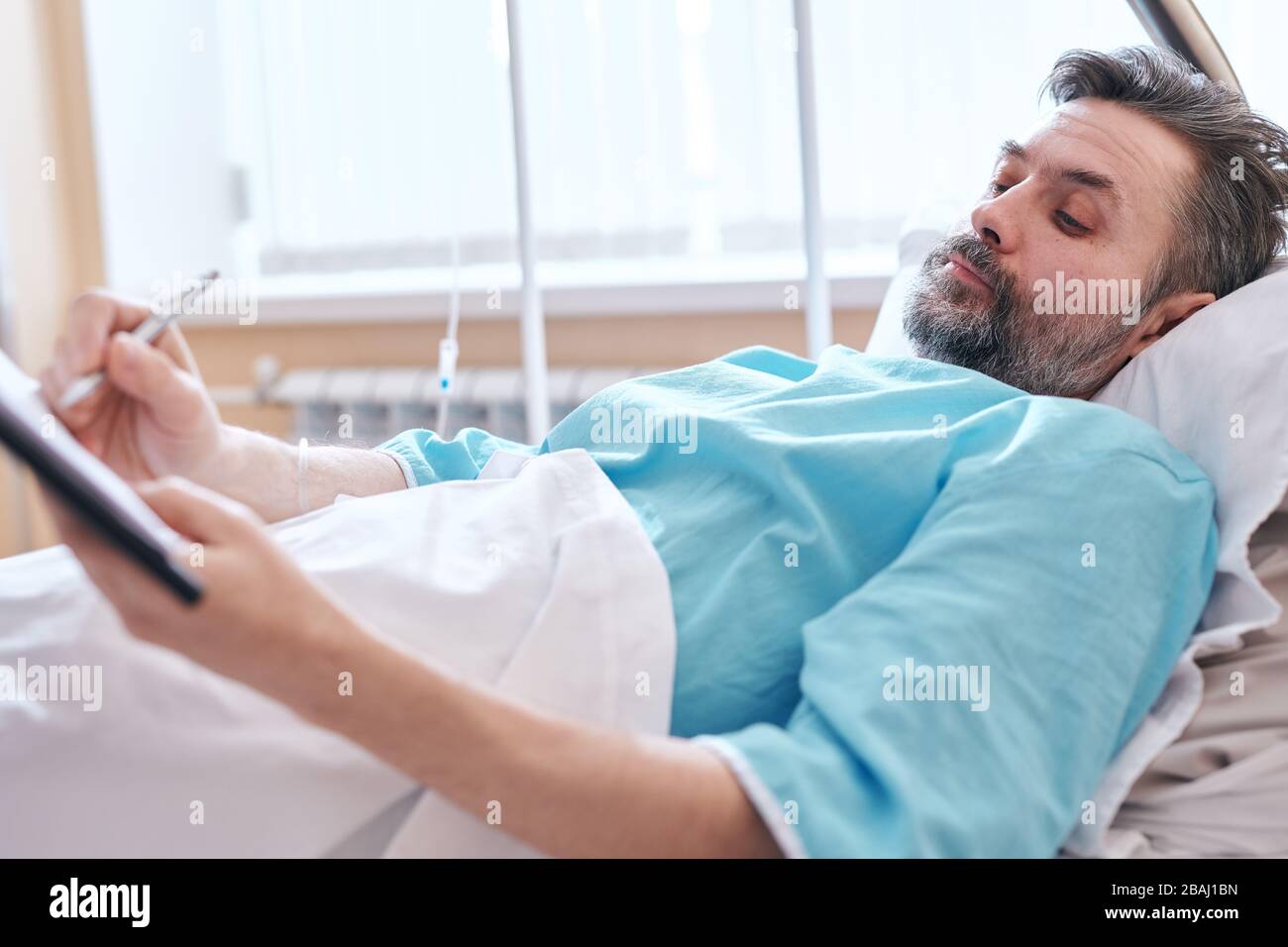 Serious gray-bearded patient lying in hospital bed and signing ...