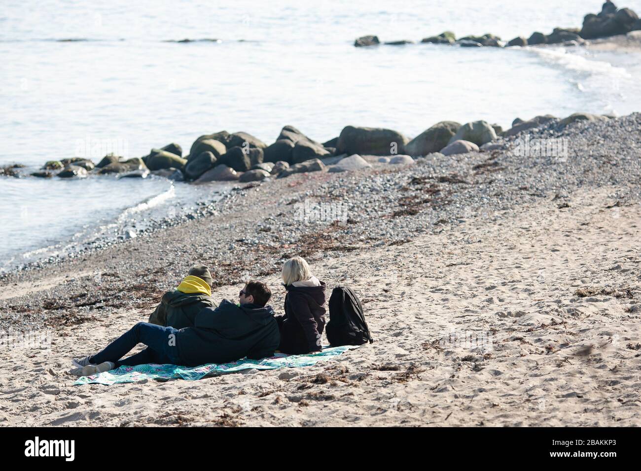 Strande, Germany. 28th Mar, 2020. Three people sit on the beach in ...