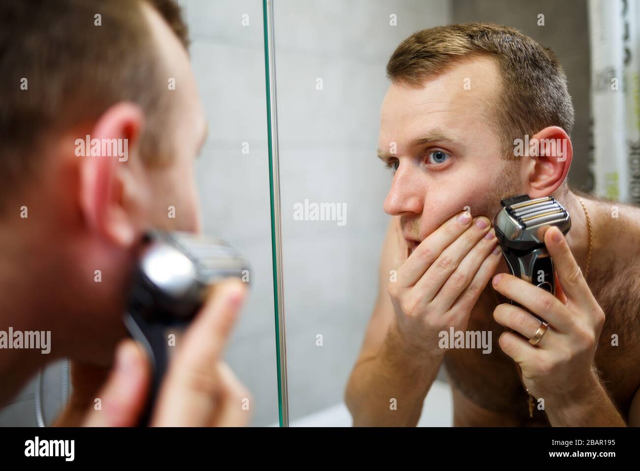 A man shaves his face with an electric razor in front of a mirror. Skin ...