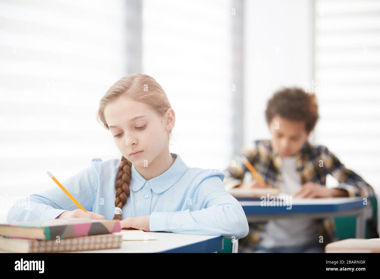 Portrait of cute blonde girl writing test while sitting at desk in ...