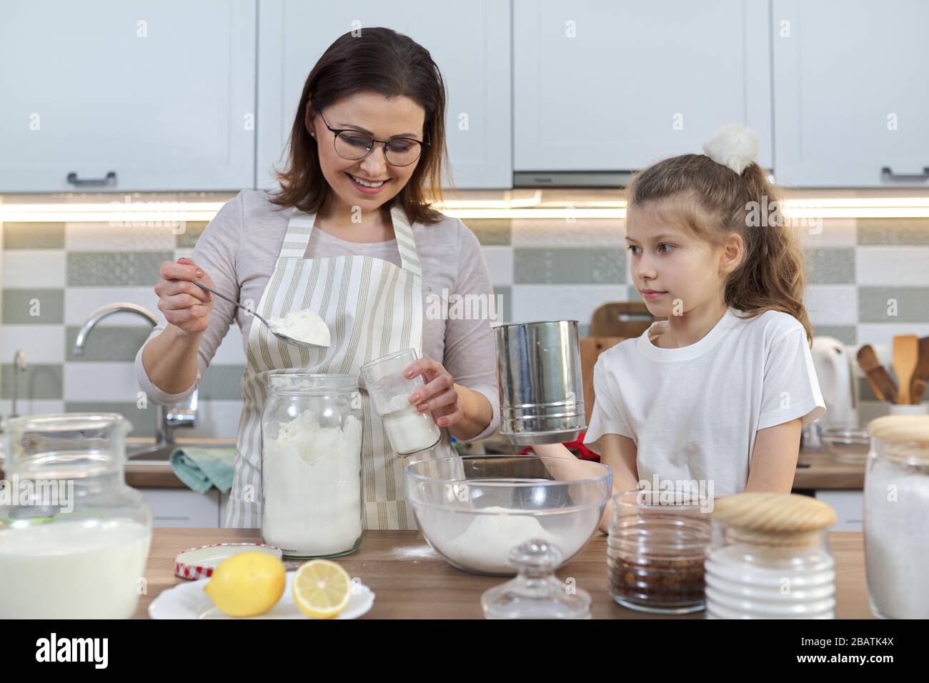 Mother and child preparing bakery together in home kitchen. Woman sifts ...