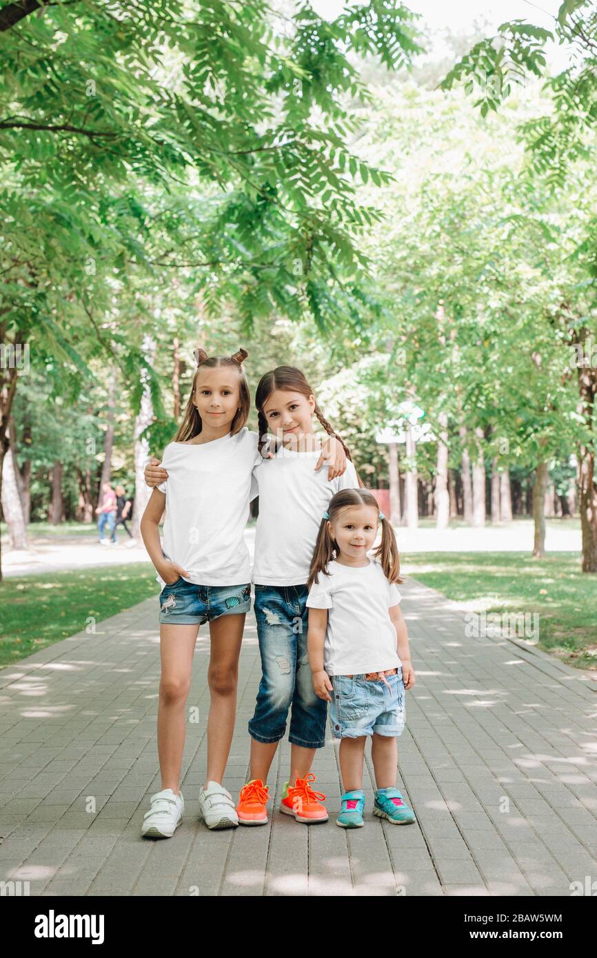Three little girls in white t-shirts stand standing and hugging in park ...
