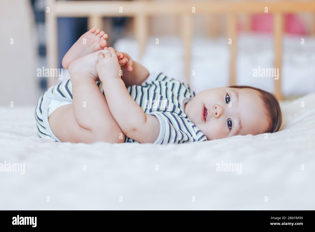 Happy baby lying on white sheet and holding her legs. Playful baby ...