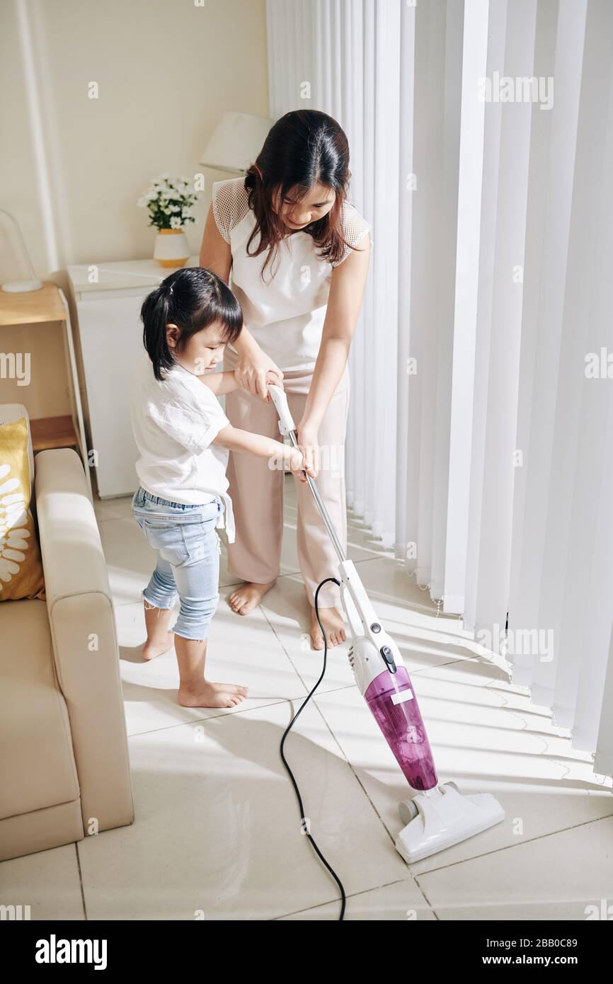 Little Asian girl helping mother with vacuum cleaning of apartment ...