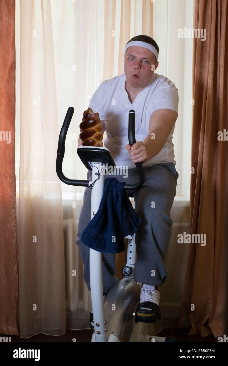 Fat guy exercising on stationary training bicycle and eating a bun at ...