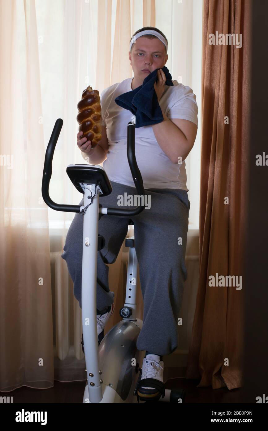 Fat guy exercising on stationary training bicycle and eating a bun at ...