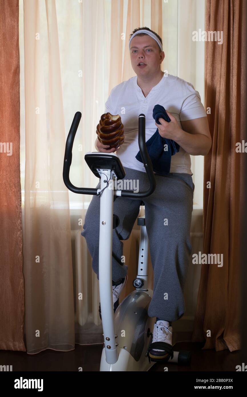 Fat guy exercising on stationary training bicycle and eating a bun at ...