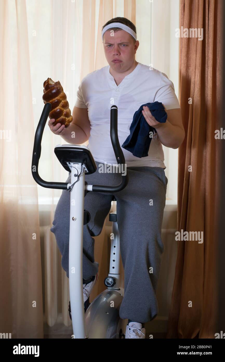 Fat guy exercising on stationary training bicycle and eating a bun at ...