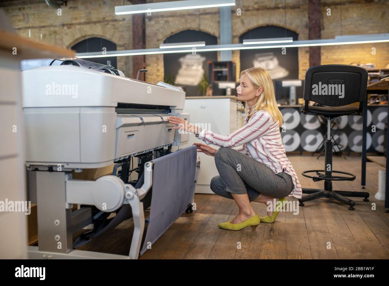 Blonde woman crouching at printing machine, checking something Stock ...
