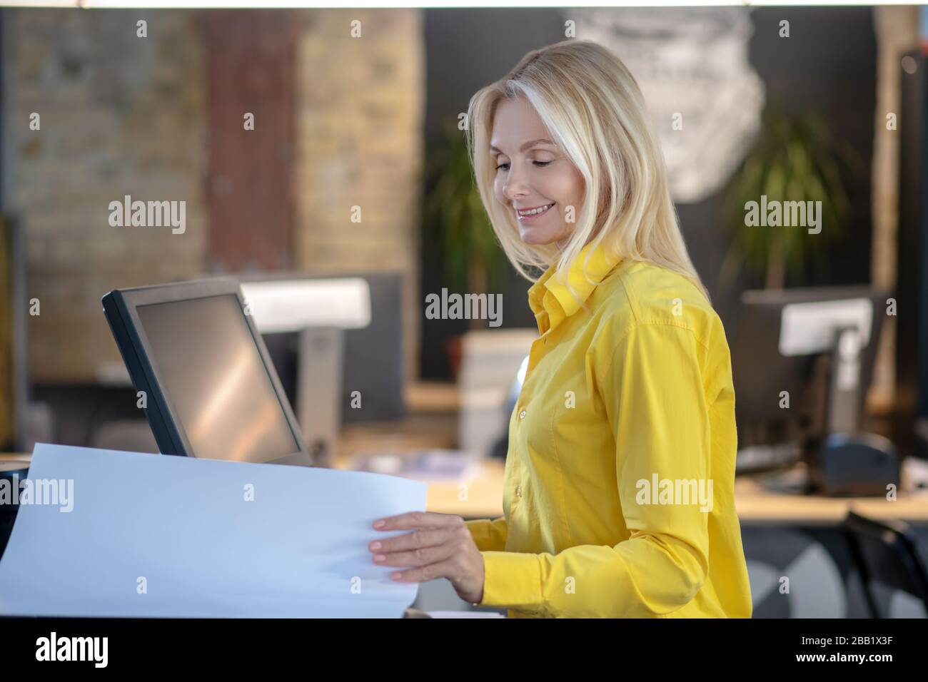 Blonde woman standing at printing machine, holding paper, smiling Stock ...