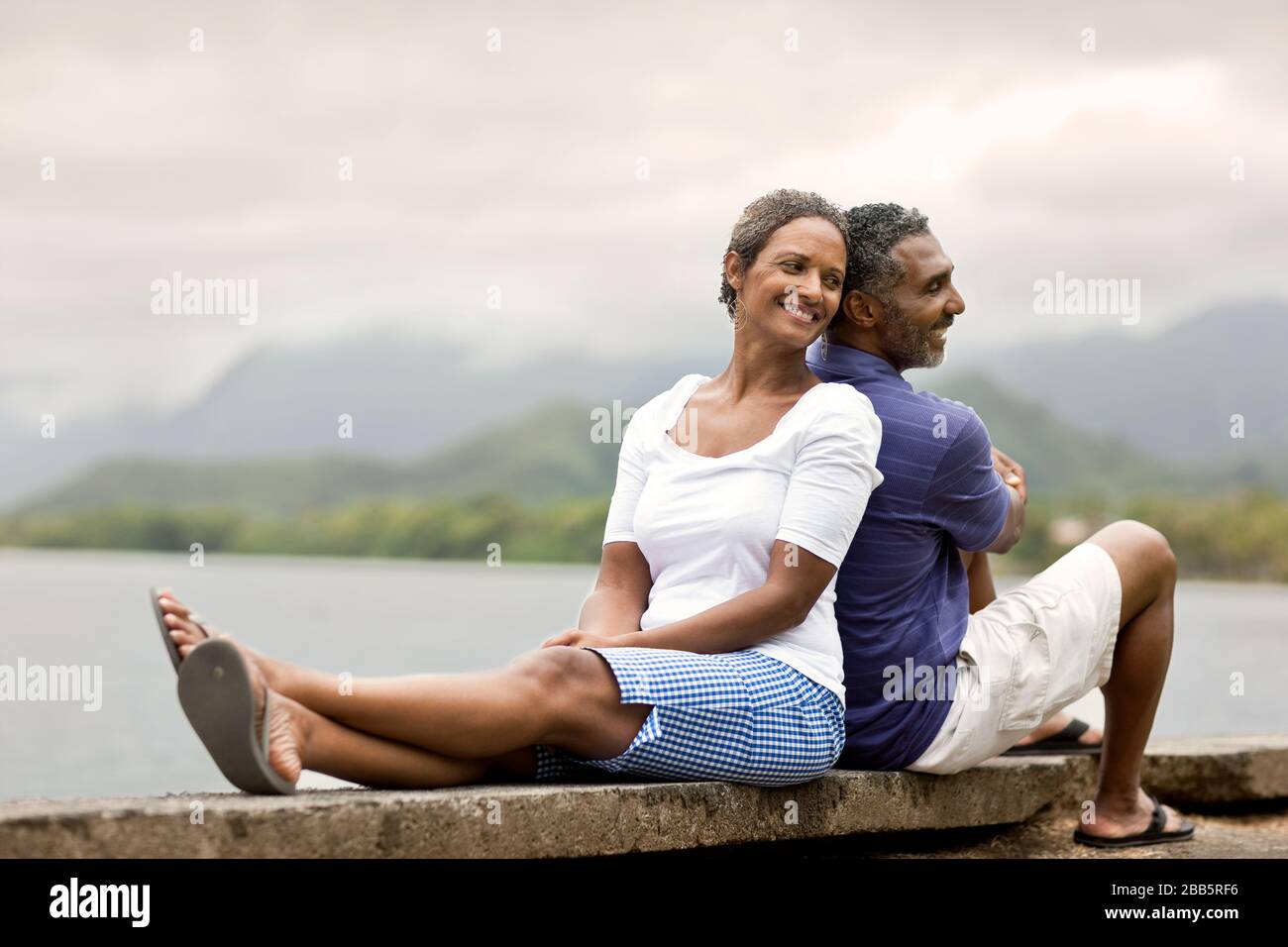 Happy mid-adult couple sitting together at the edge of a waterfront ...