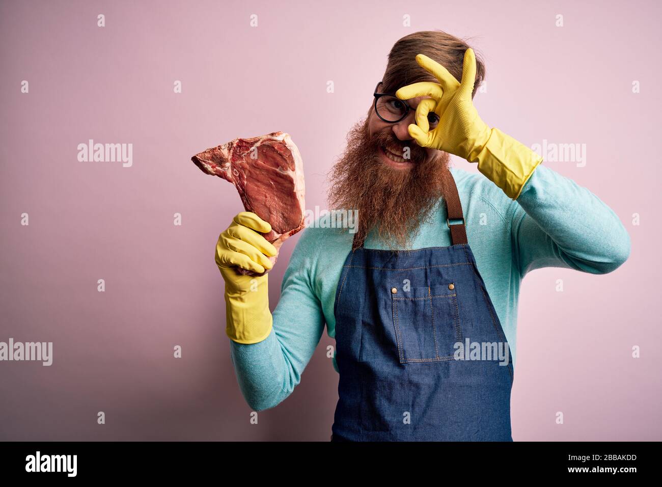 Redhead Irish butcher man with beard holding raw beef steak over pink ...