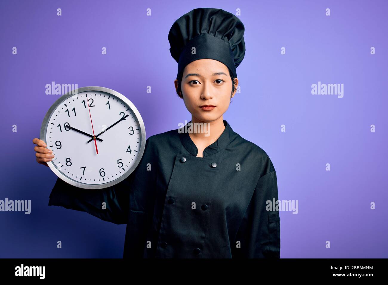 Young chinese chef woman wearing cooker uniform and hat holding clock ...