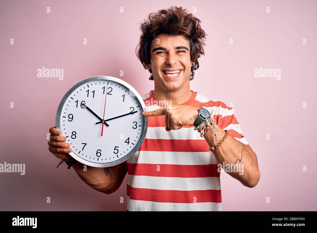 Young handsome man holding big clock standing over isolated pink ...