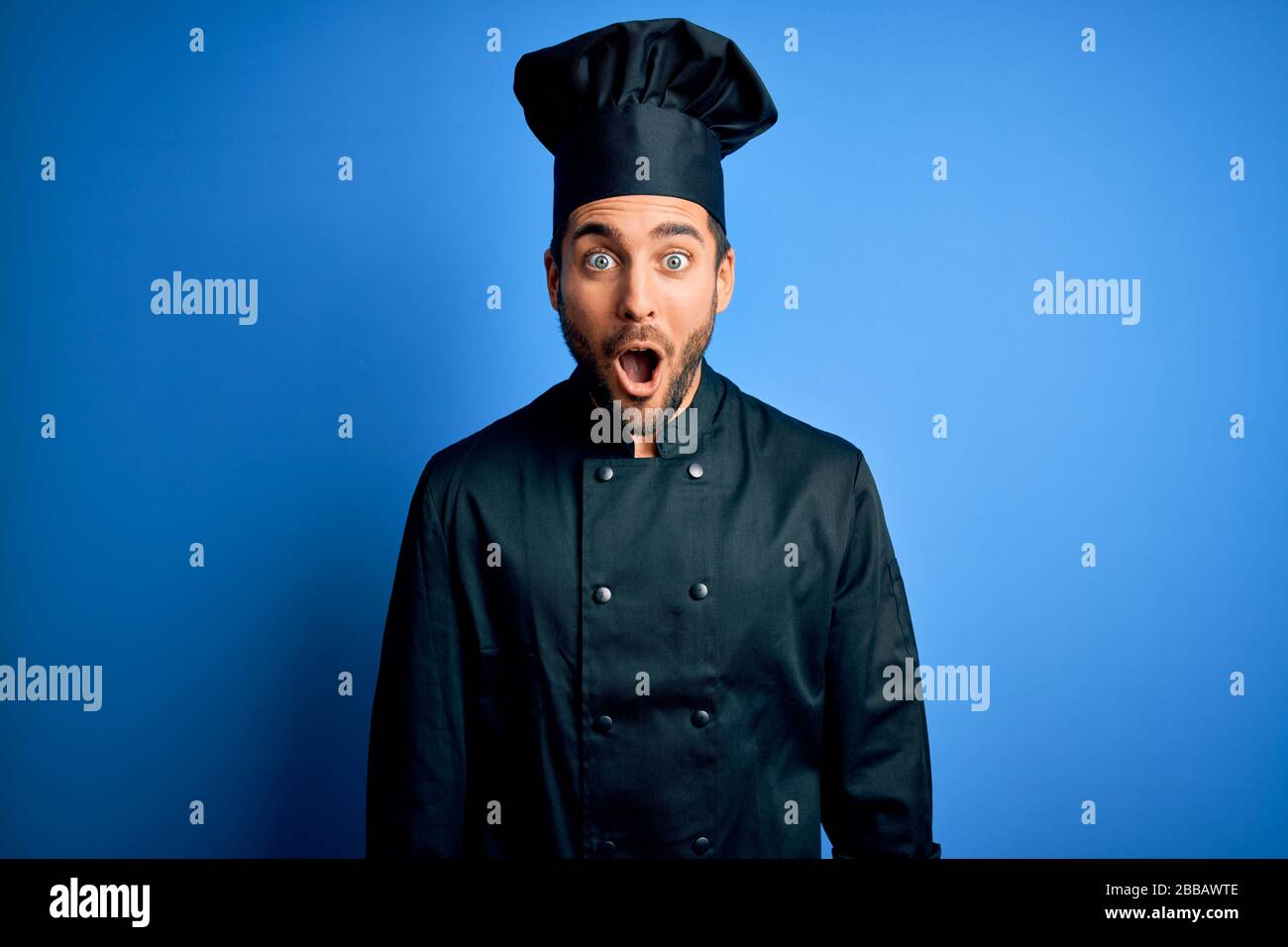 Young handsome chef man with beard wearing cooker uniform and hat over ...