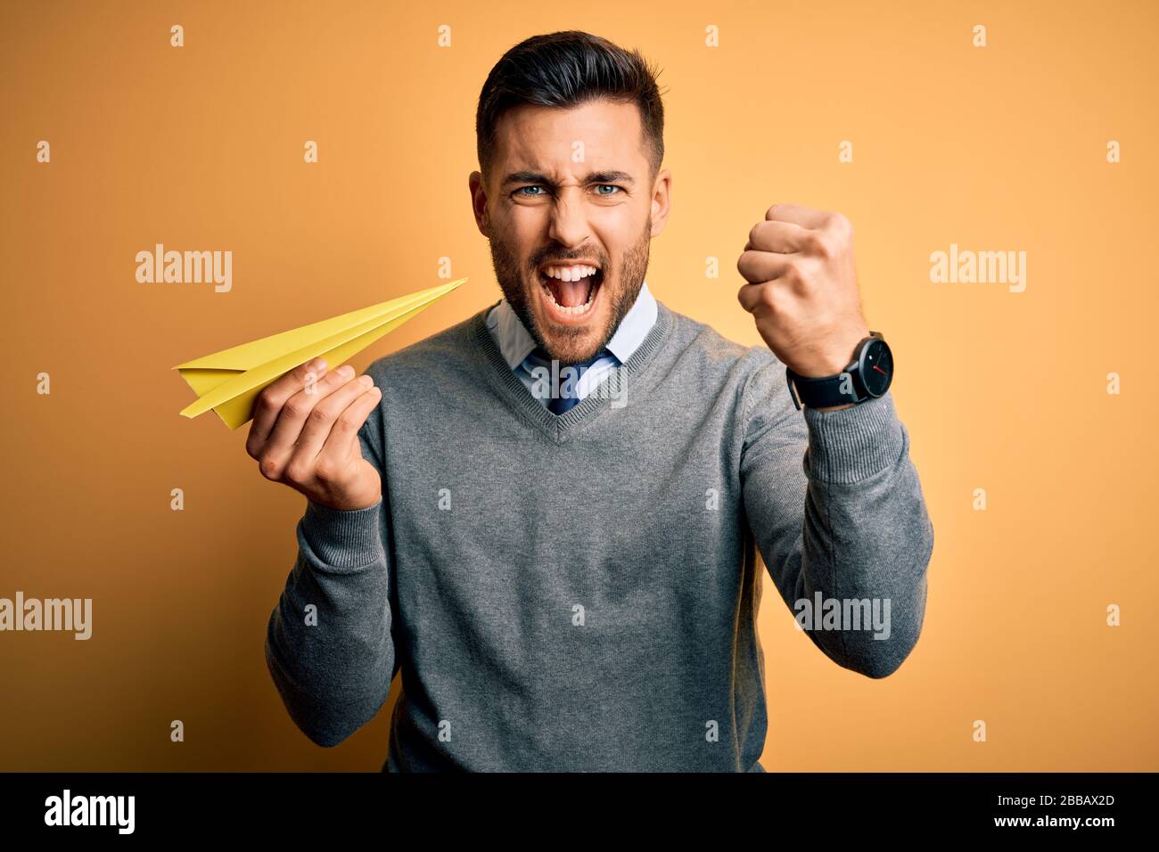 Young handsome man holding paper airplane standing over isolated yellow ...
