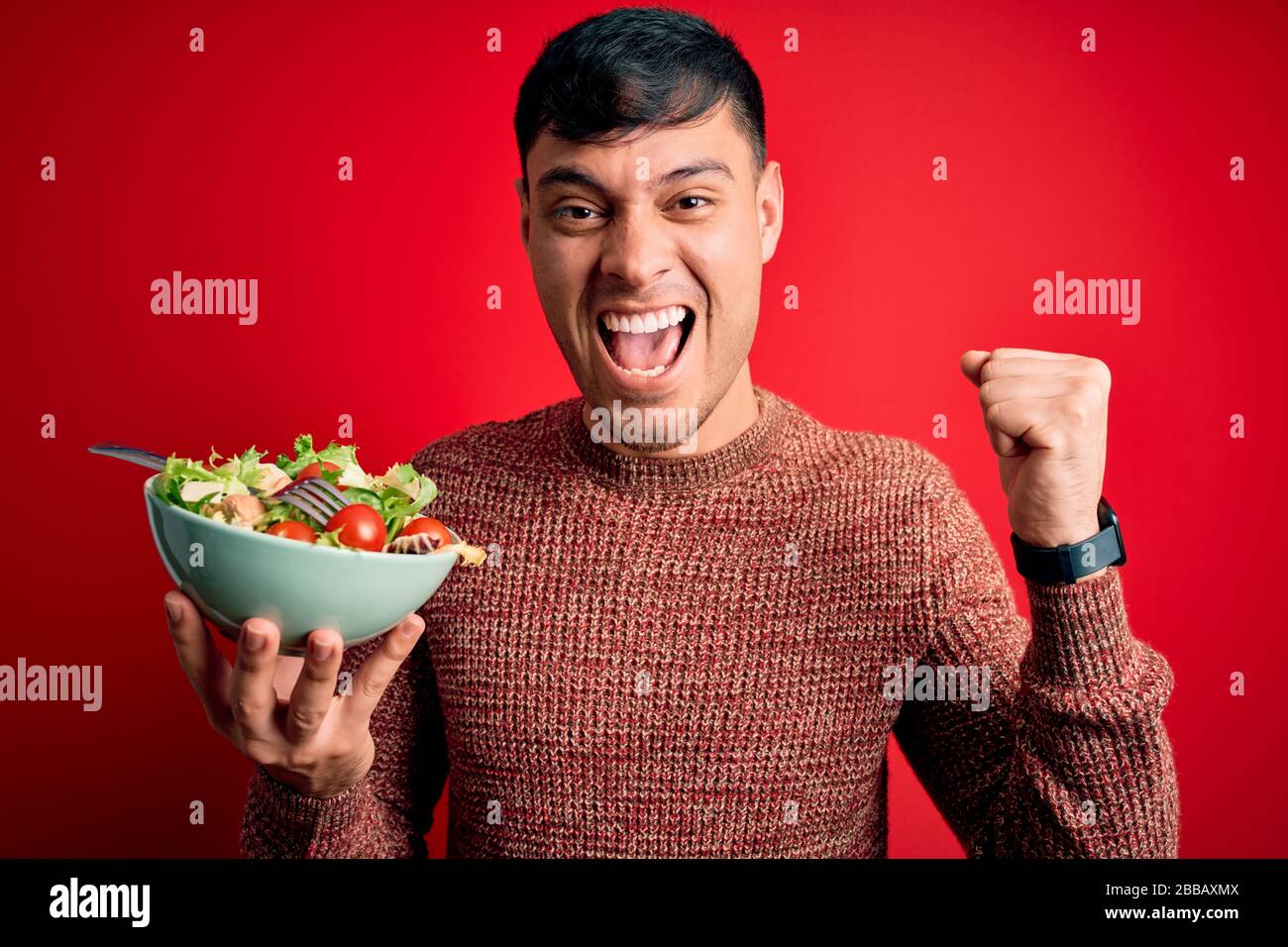 Young hispanic man eating vegetarian healthy salad over red isolated ...