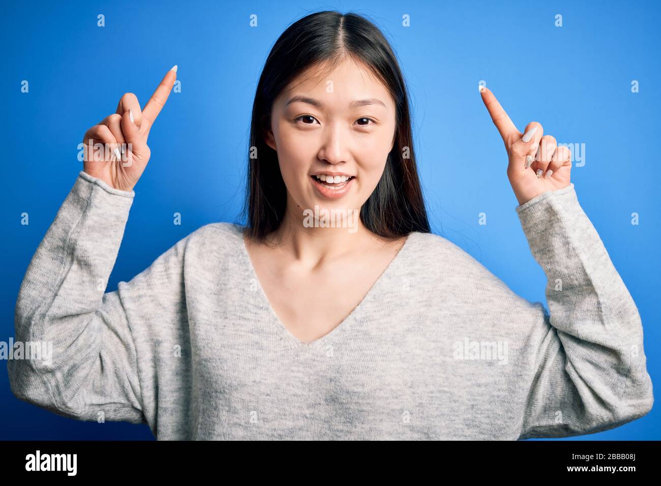 Young beautiful asian woman wearing casual sweater standing over blue ...