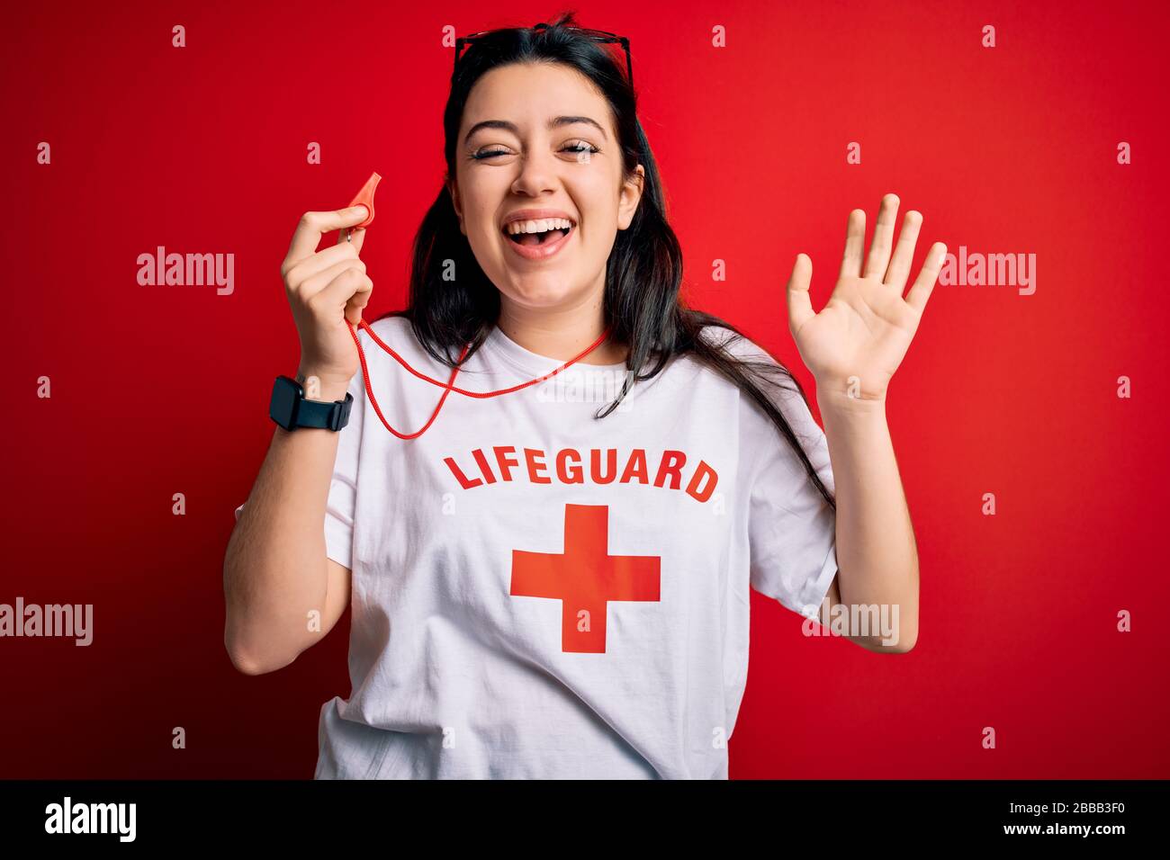 Young lifeguard woman wearing guard equipement holding whistle over red ...