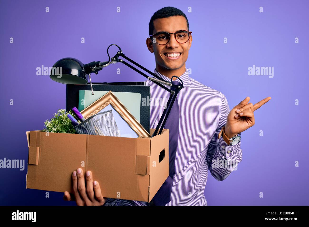 Handsome african american man fired holding box with work objects over purple background very ...