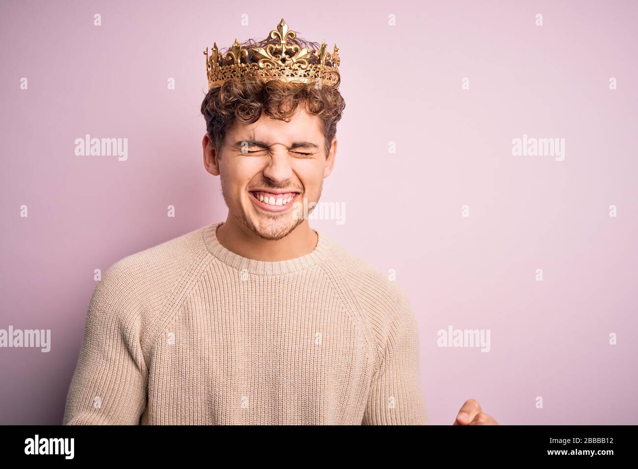 Young blond man with curly hair wearing golden crown of king over pink ...