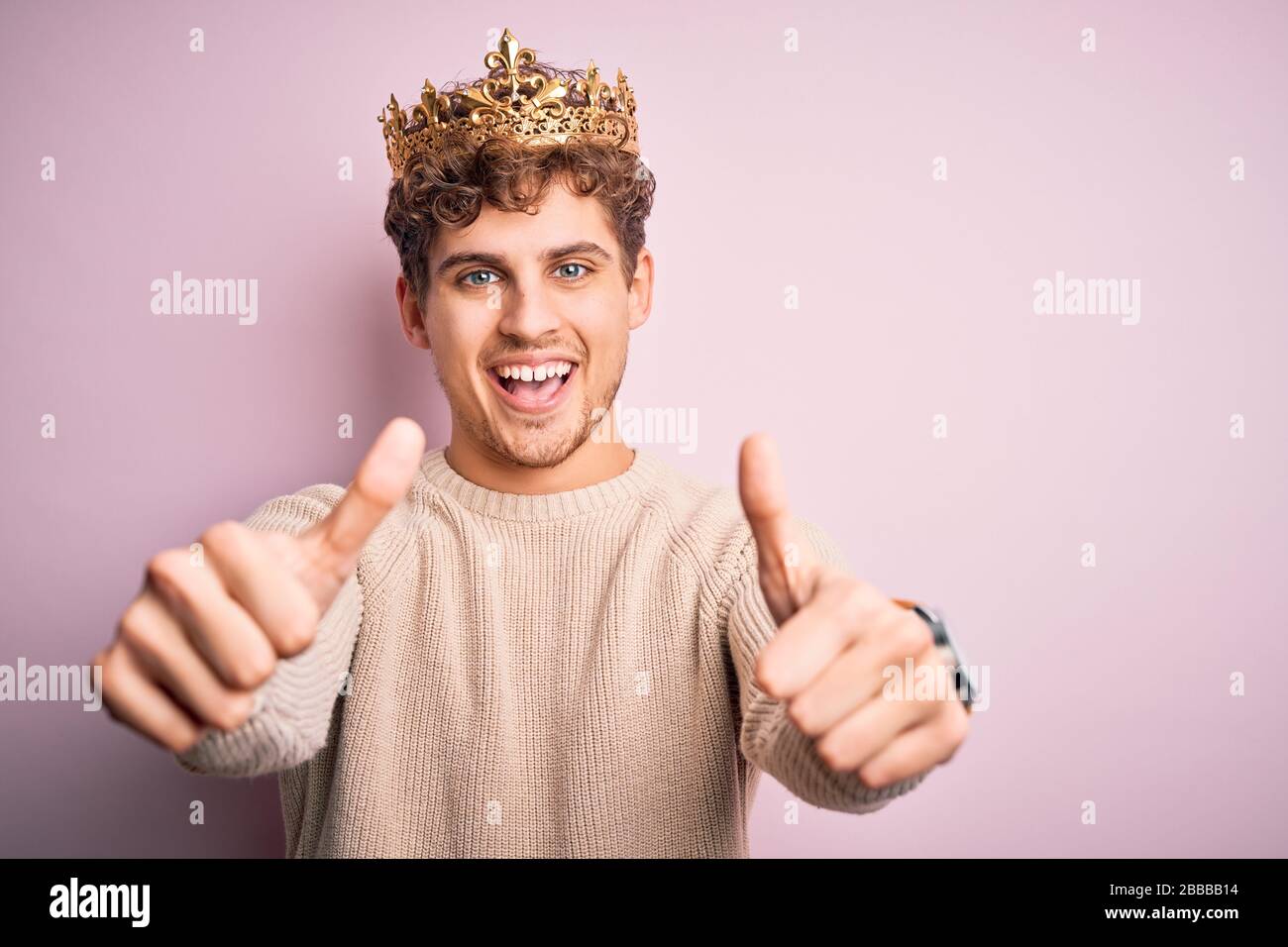 Young blond man with curly hair wearing golden crown of king over pink ...
