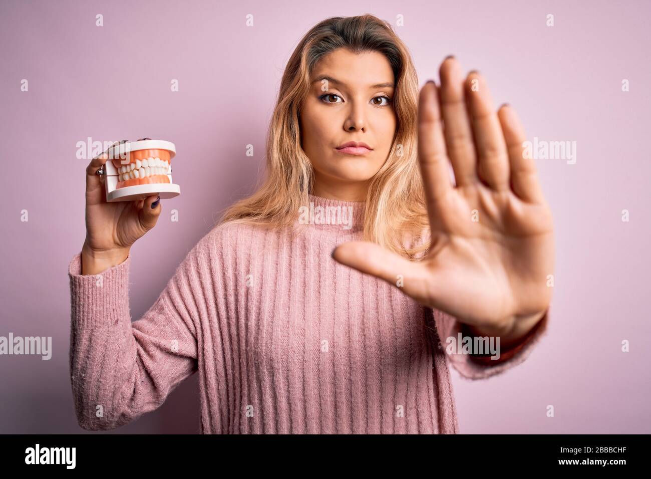 Young beautiful blonde woman holding plastic denture over isolated pink ...