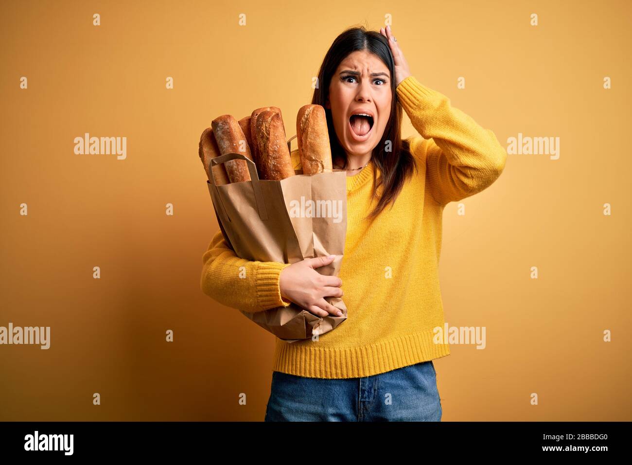 Young beautiful woman holding a bag of fresh healthy bread over yellow ...