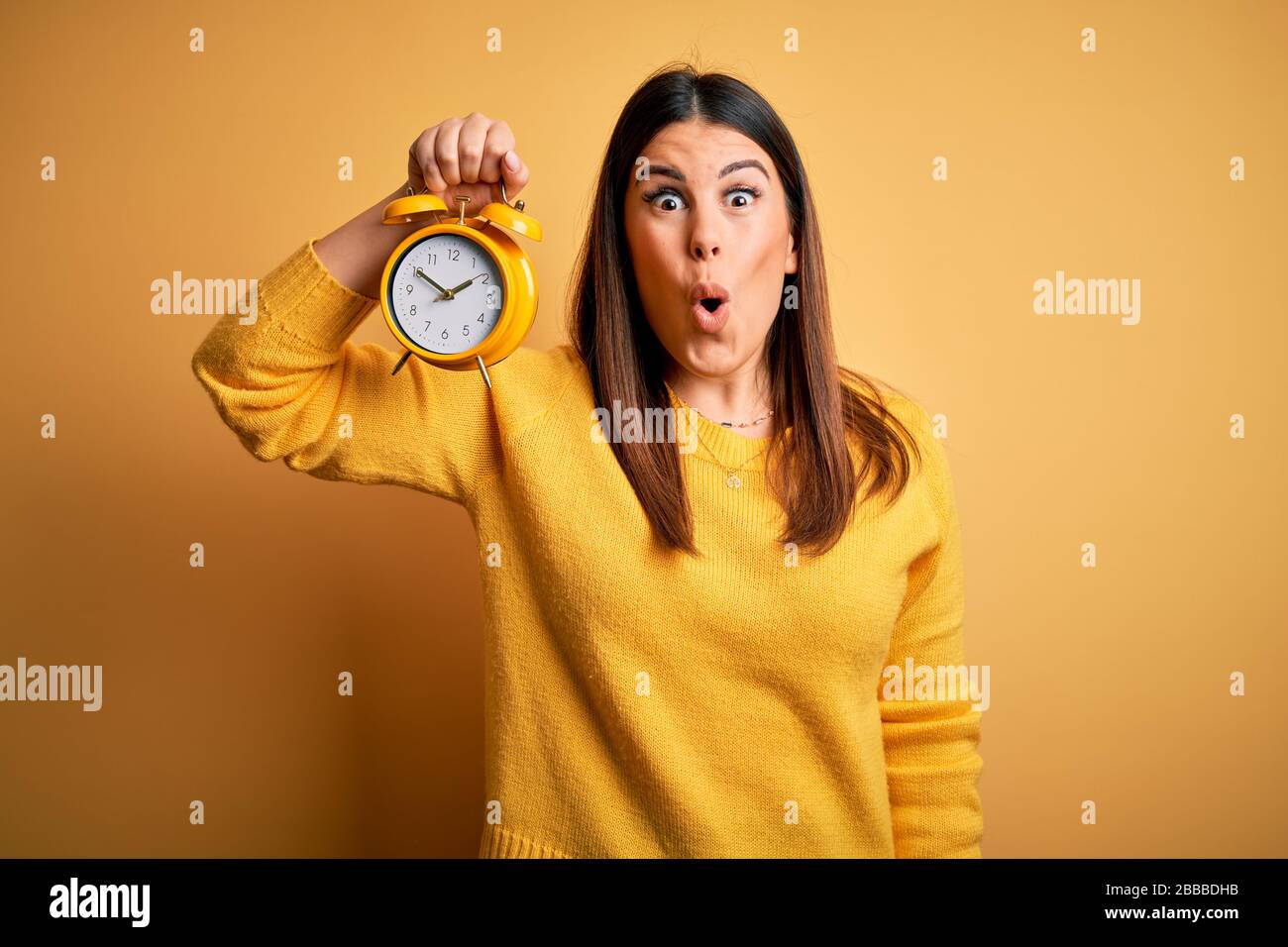 Young beautiful woman holding alarm clock standing over isolated yellow ...