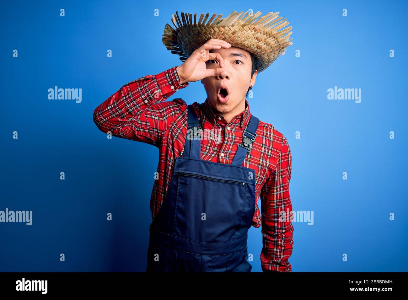 Young handsome chinese farmer man wearing apron and straw hat over blue ...