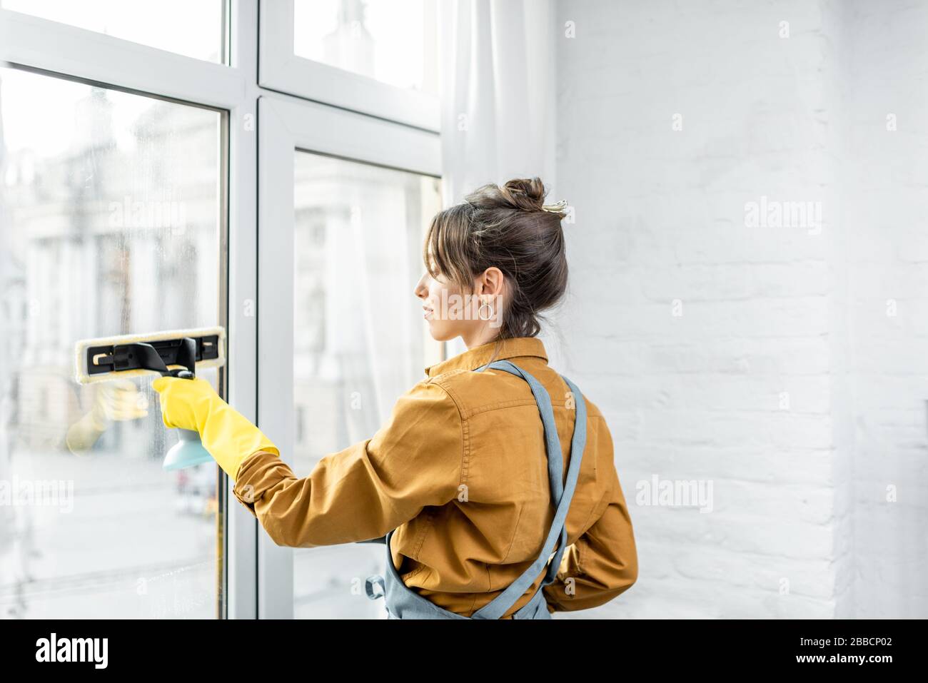 Beautiful young housewife in working clothes washing windows by ...
