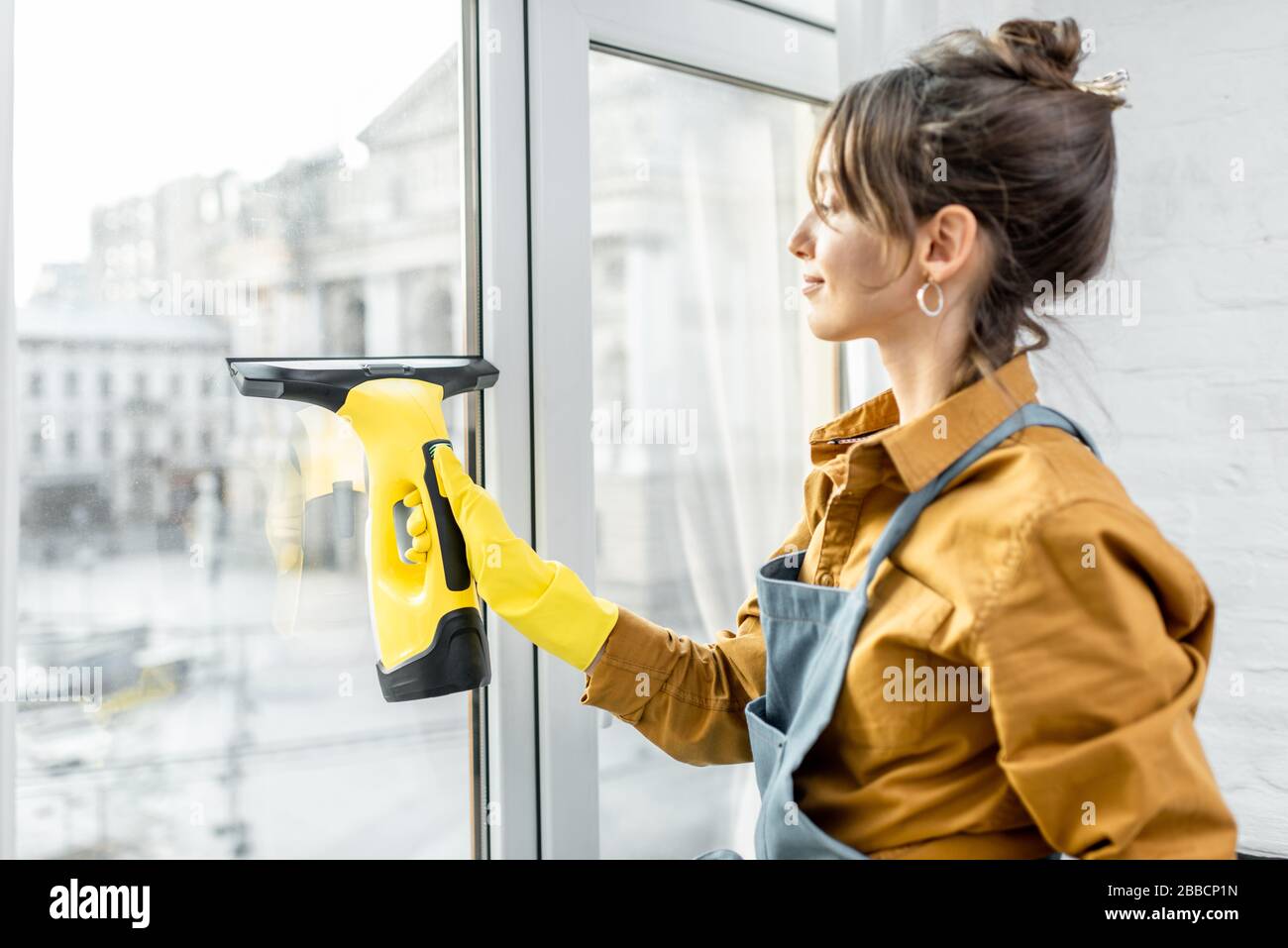 Beautiful young housewife in working clothes washing windows by ...