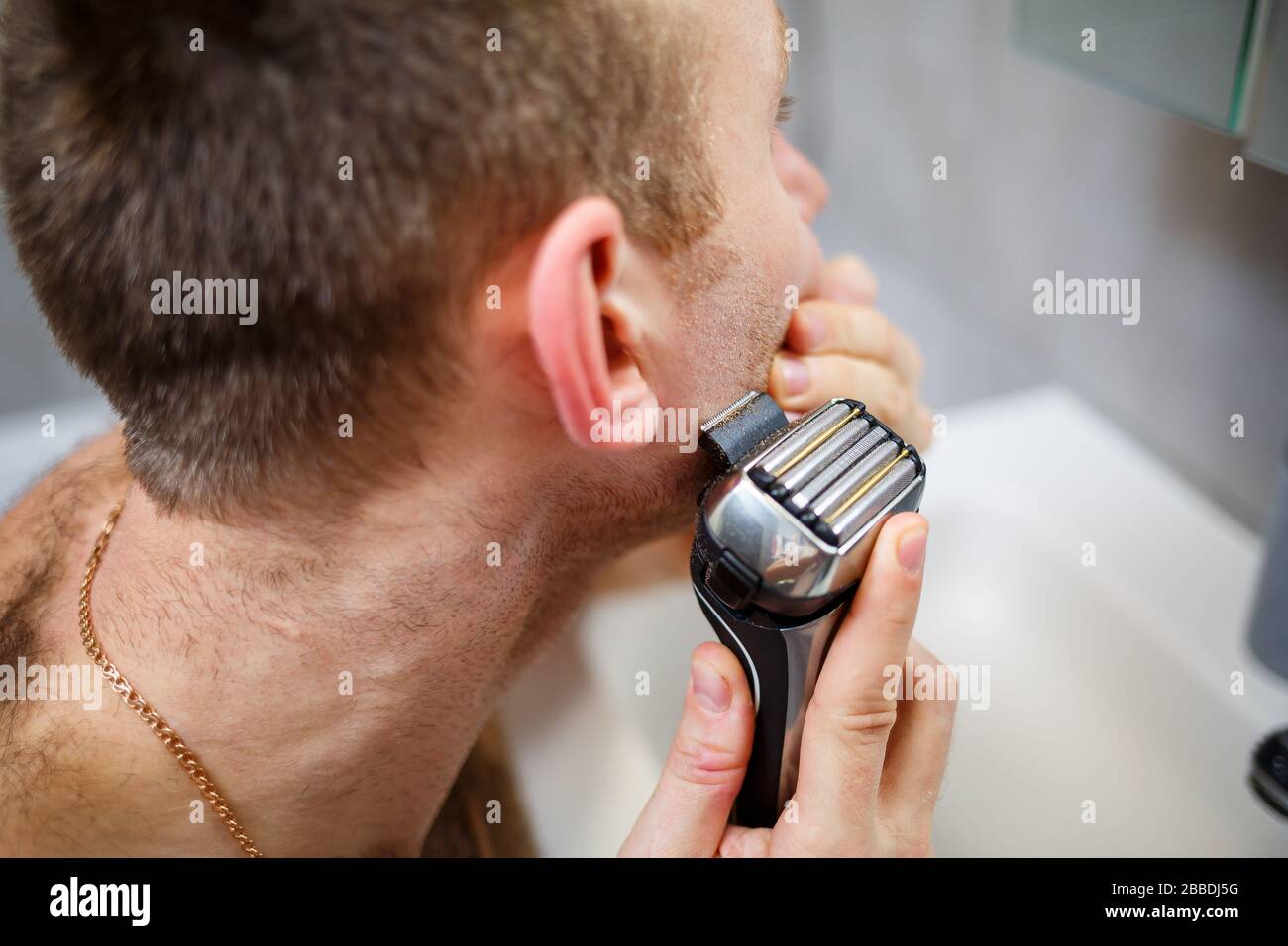 A man shaves his face with an electric razor in front of a mirror. Skin ...