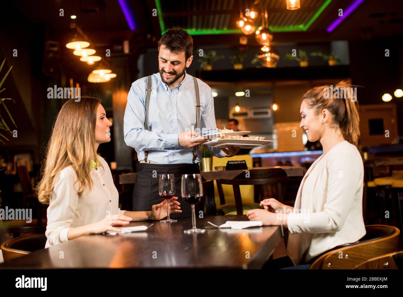 Two smiling young female friends at a restaurant with waiter serving ...