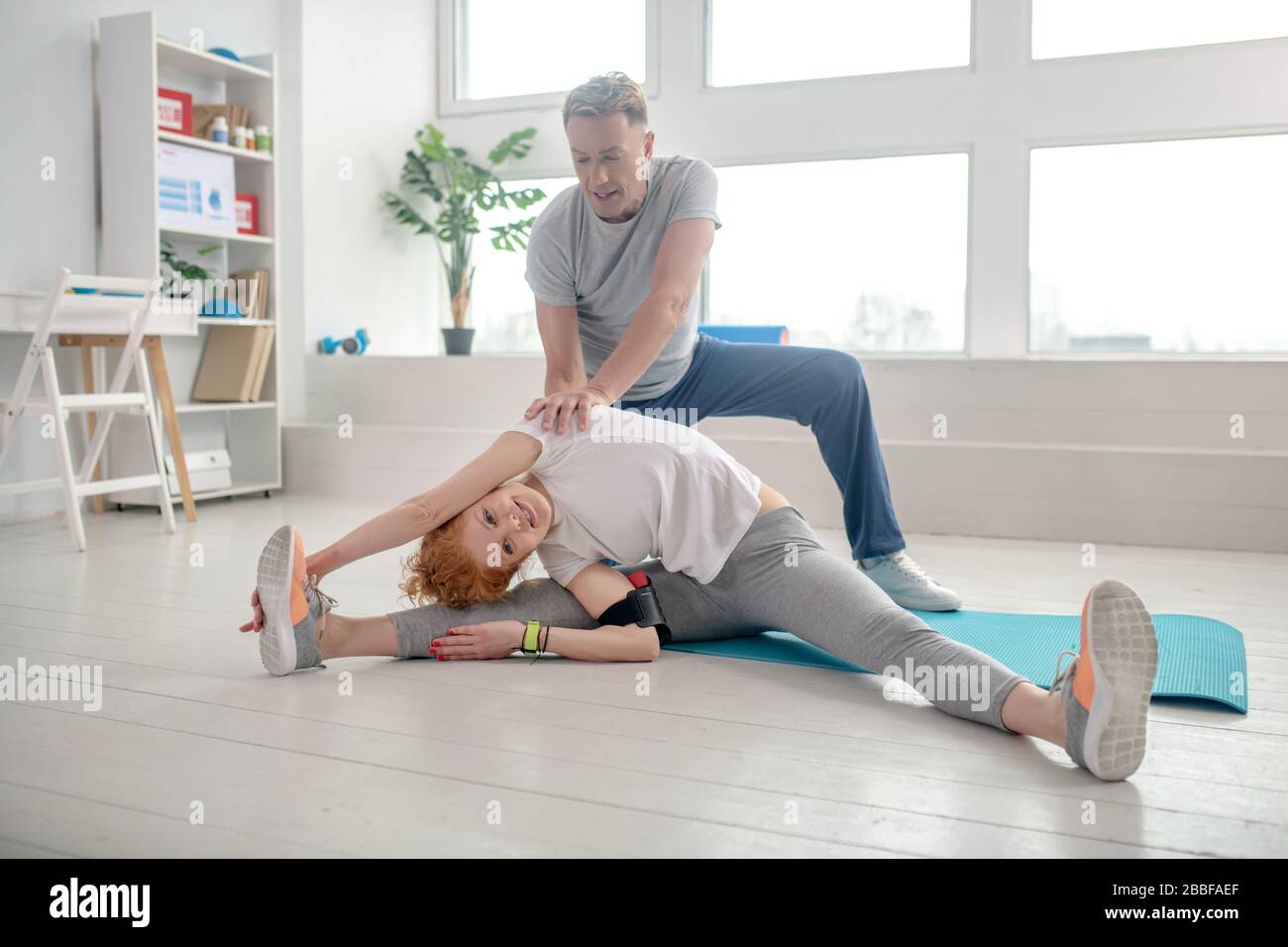 Female patient practicing straddle splits on mat, male physiotherapist ...