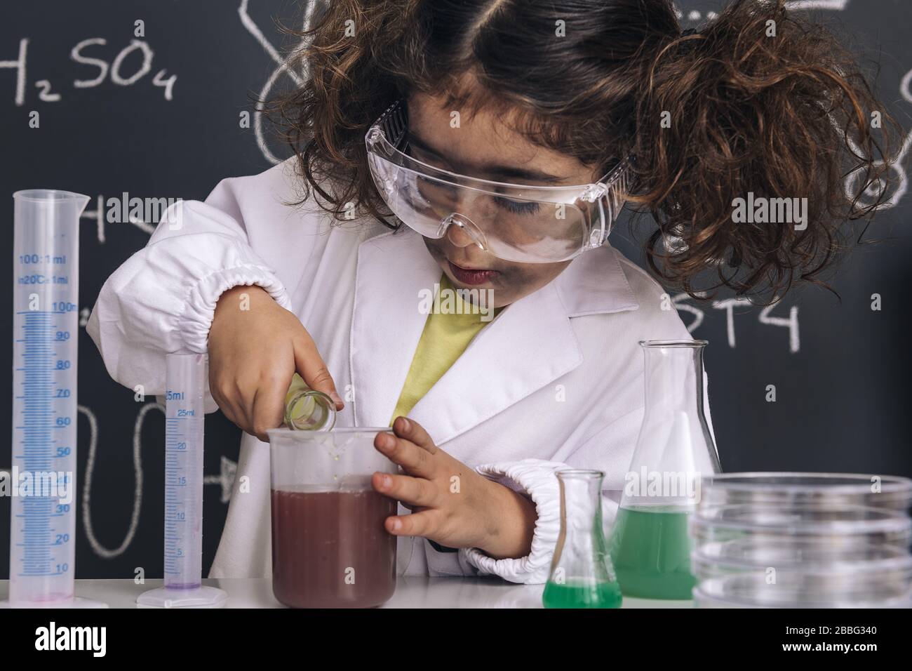 focused scientist child with goggles and gloves in lab coat mixing ...
