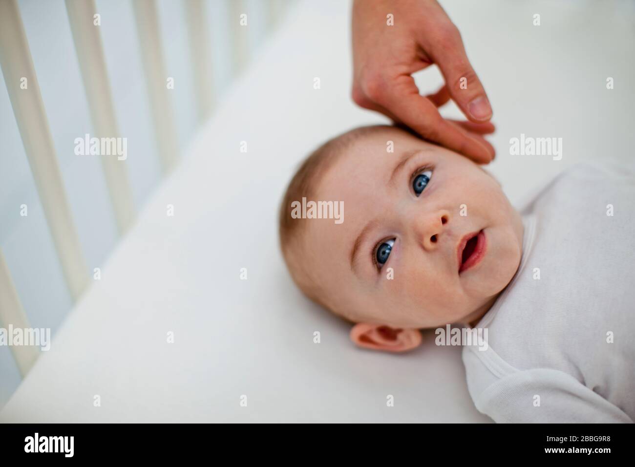 Cute baby boy gazing up out of his crib as his young mother gently ...