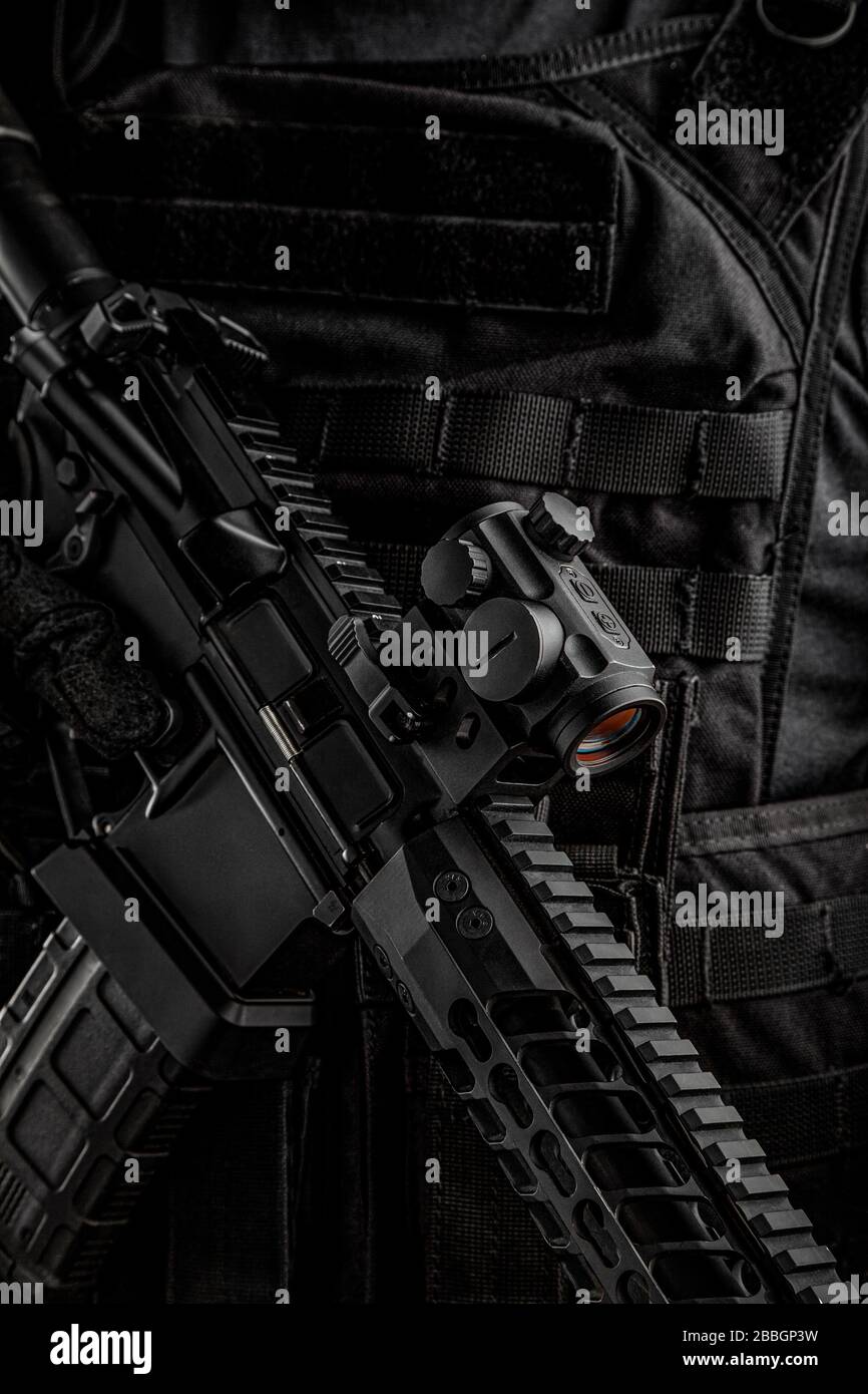 A man holds an automatic carbine with a collimator sight on a dark ...