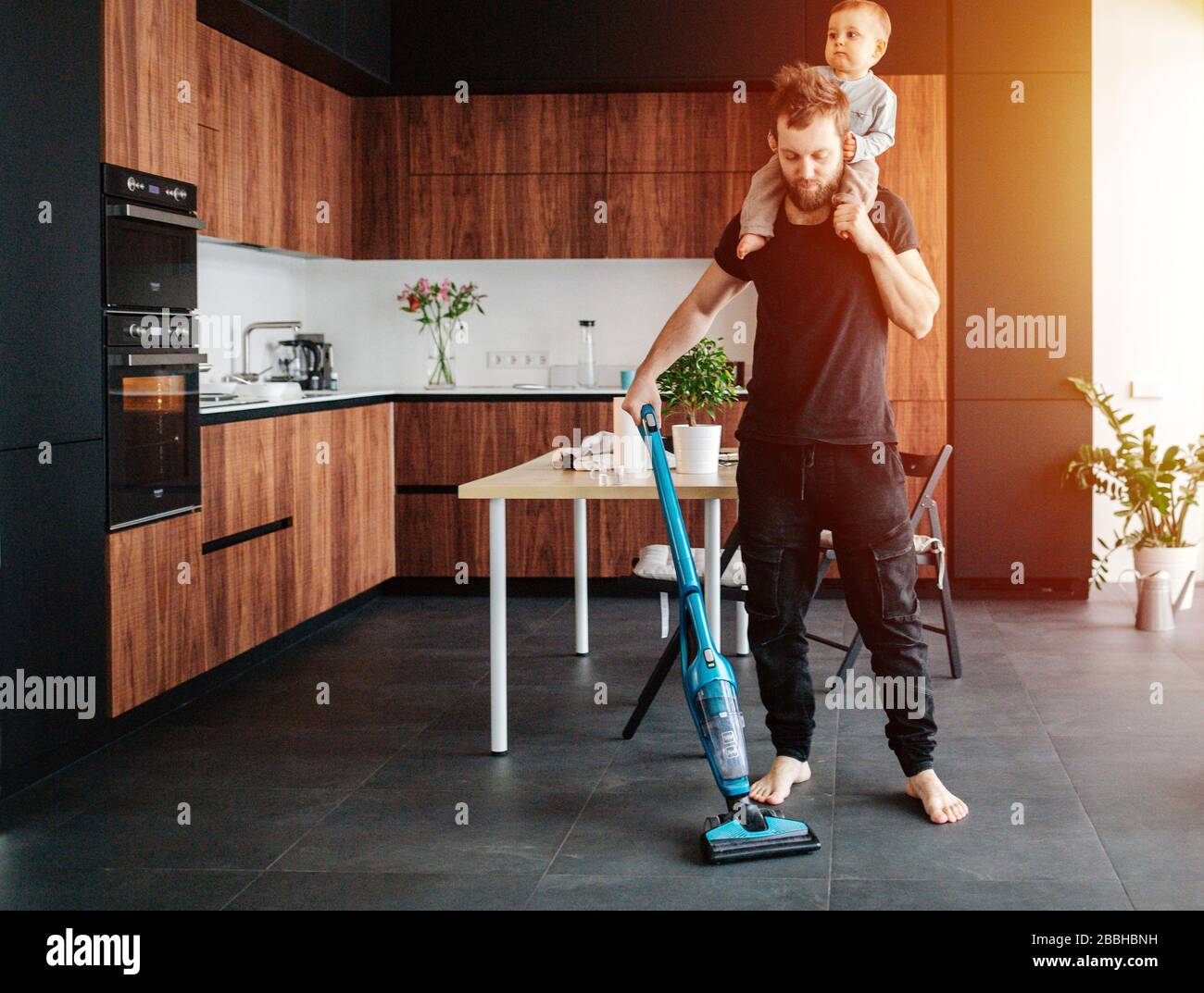 Young father vacuums apartment floor with his baby riding on his neck ...